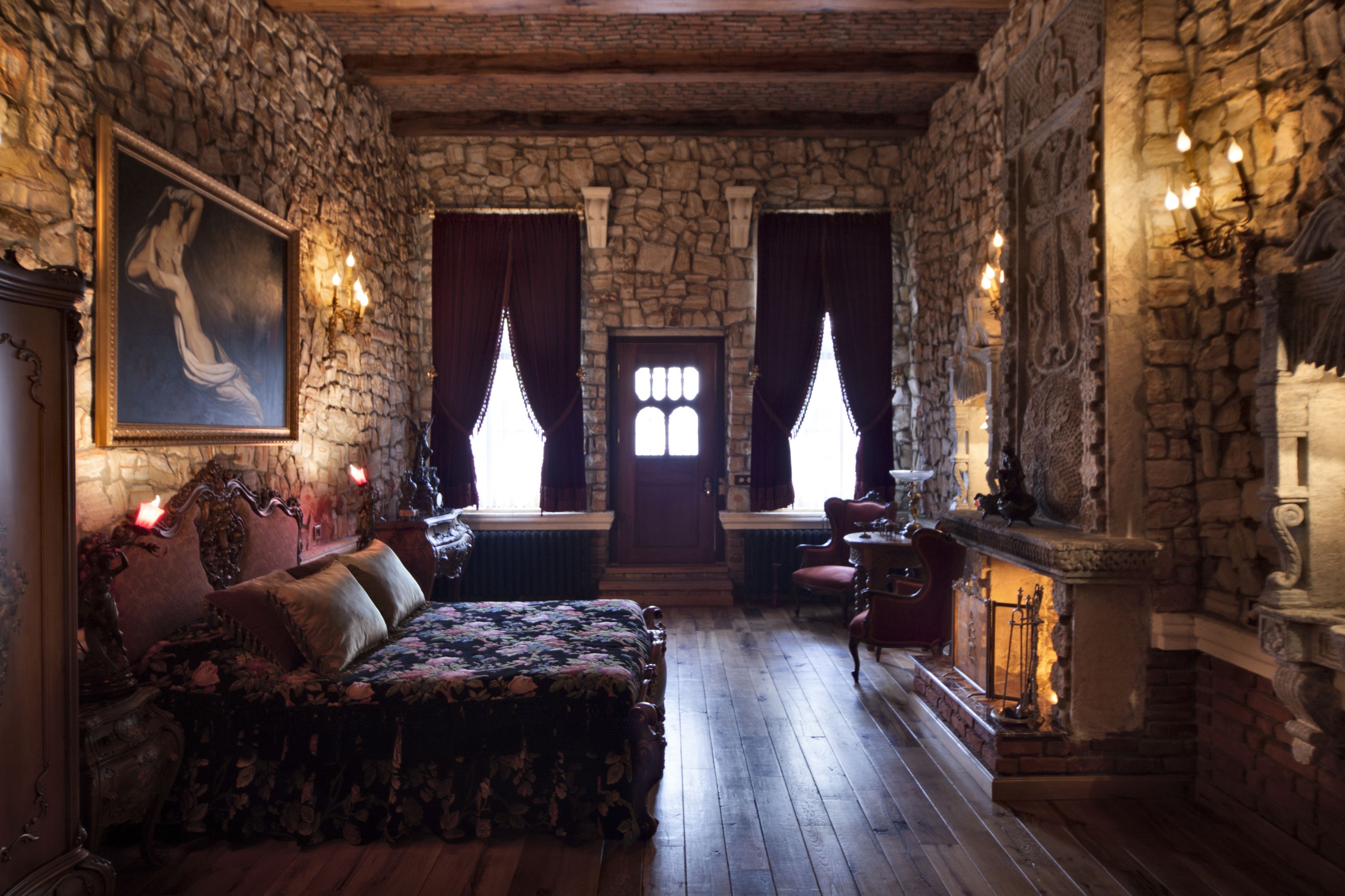 Quarto com paredes de pedra, cama com roupa de cama floral, lareira e um quadro na parede.