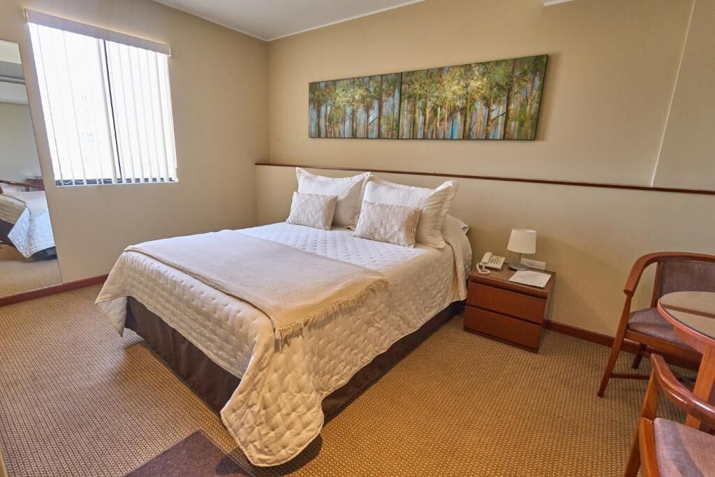 Quarto de hotel com uma cama grande, mesa de cabeceira com abajur, uma cadeira e um quadro na parede.