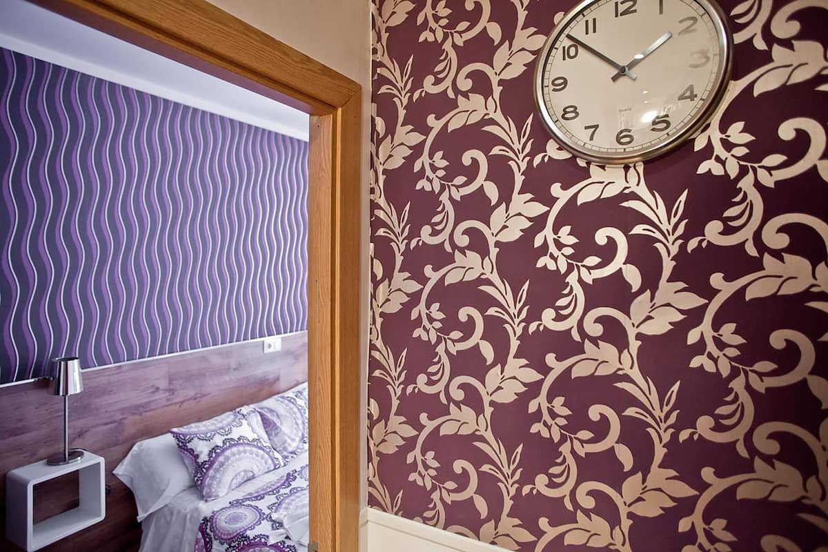 Quarto com papel de parede estampado, um relógio, uma cama com roupa de cama estampada e uma mesa de cabeceira com abajur.
