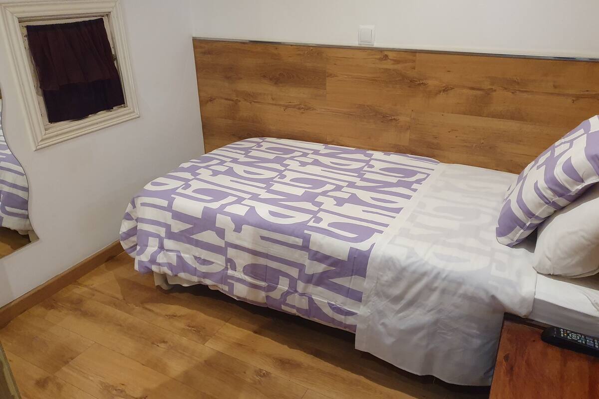 Quarto com cabeceira de madeira, cama com colcha estampada, mesa de cabeceira com controle remoto e uma pequena janela com cortina.