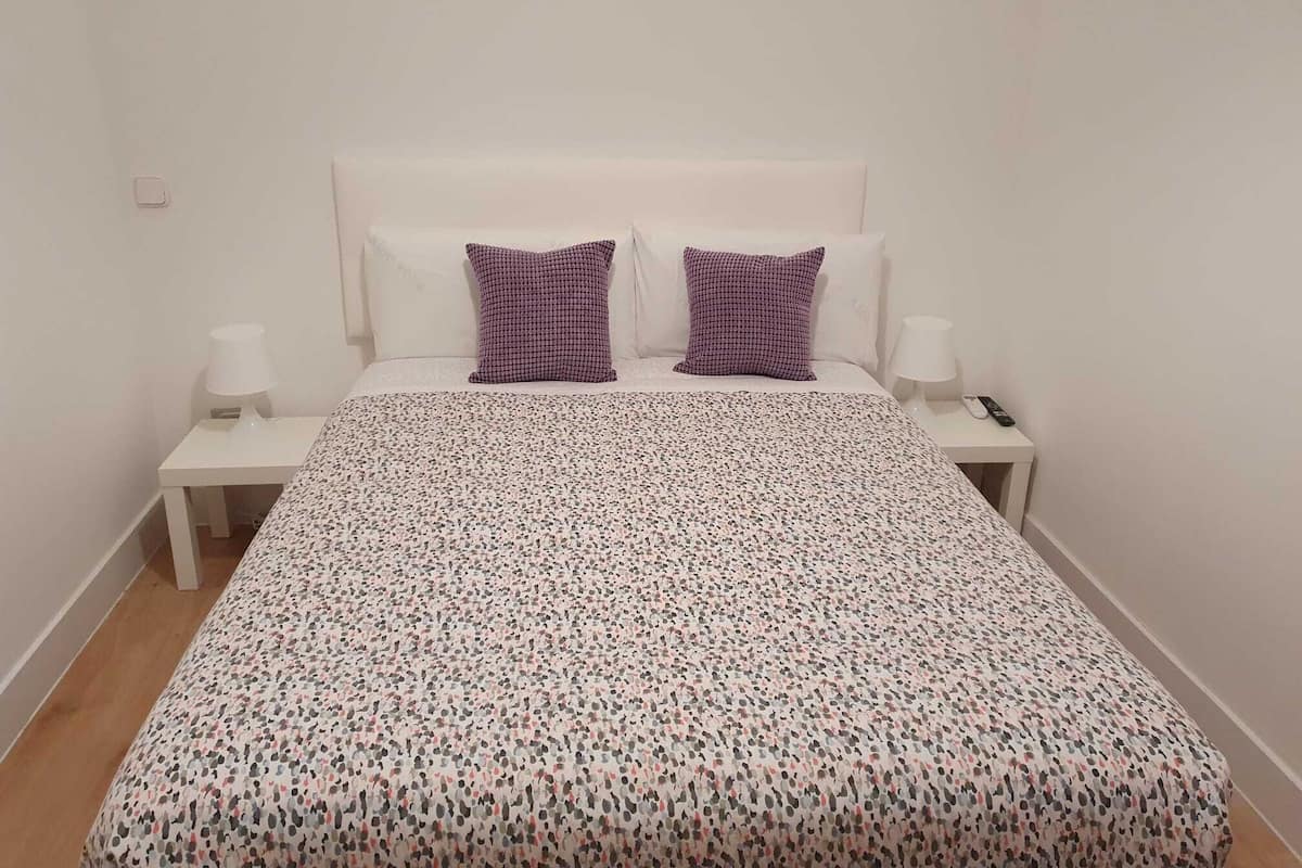 Uma cama bem arrumada com colcha estampada, duas almofadas e duas luminárias de cabeceira sobre mesas de cabeceira brancas.