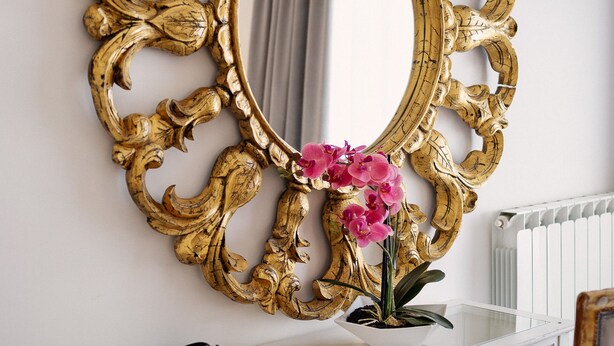 Quarto com um espelho grande e ornamentado em moldura dourada, uma mesa consola branca com gavetas, uma câmera e uma cadeira.