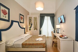 Quarto de hotel com cama, mesa de cabeceira, escrivaninha e televisão.