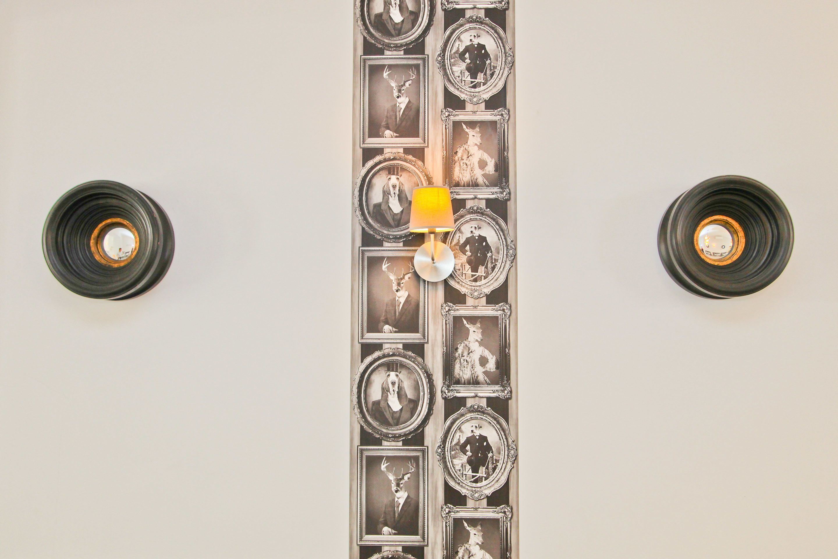 Paredes com quadros emoldurados dispostos em linha vertical, uma luminária no centro e dois objetos circulares em cada lado.