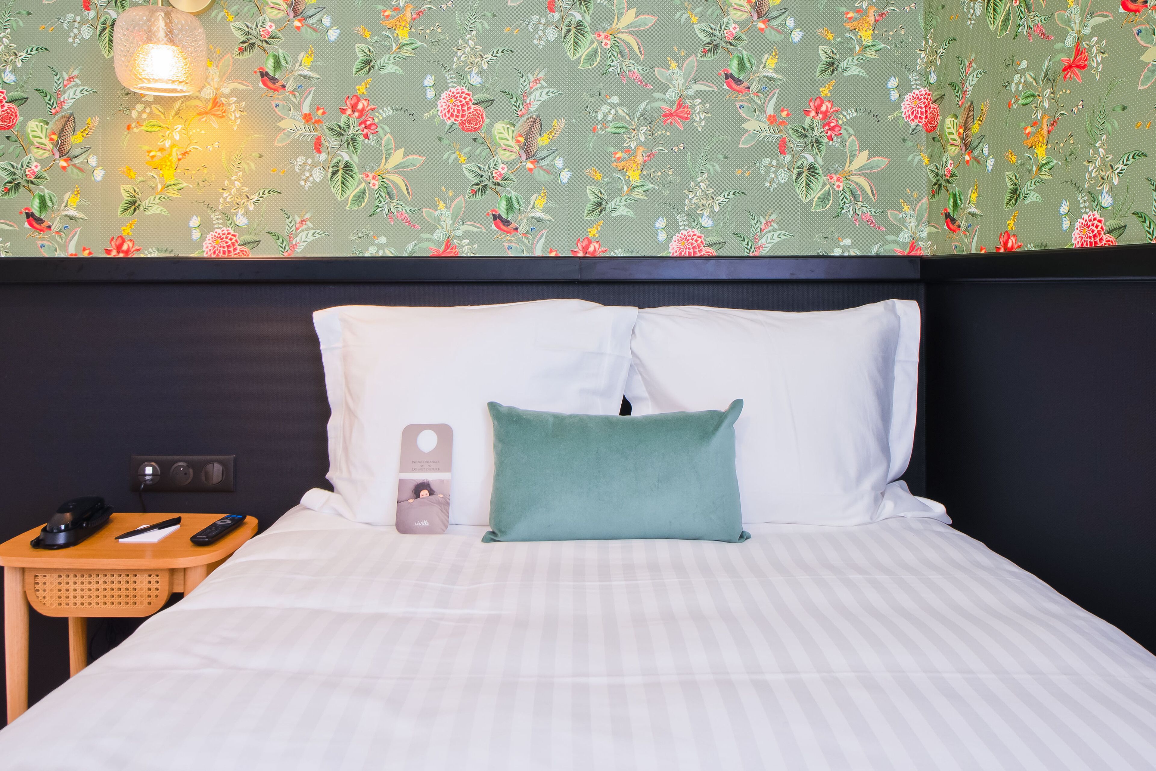 Cama bem arrumada com almofadas brancas e verdes, mesa de cabeceira com telefone e controle remoto, e papel de parede com padrão floral na parede.