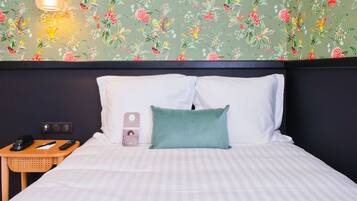 Cama bem arrumada com almofadas brancas e verdes, mesa de cabeceira com telefone e controle remoto, e papel de parede com padrão floral na parede.