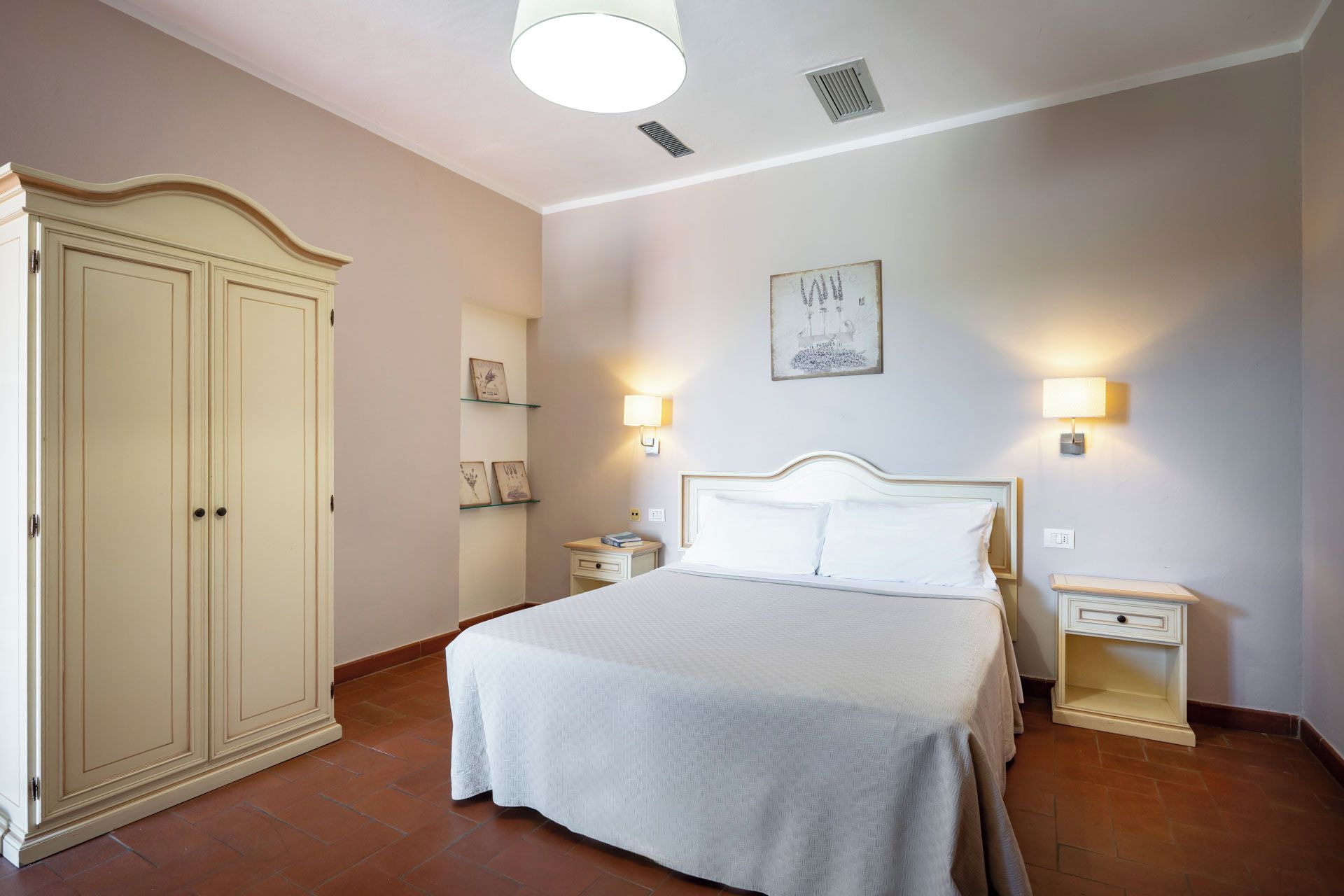 Quarto com cama, mesas de cabeceira, guarda-roupa, luminárias na parede e um respiradouro no teto.