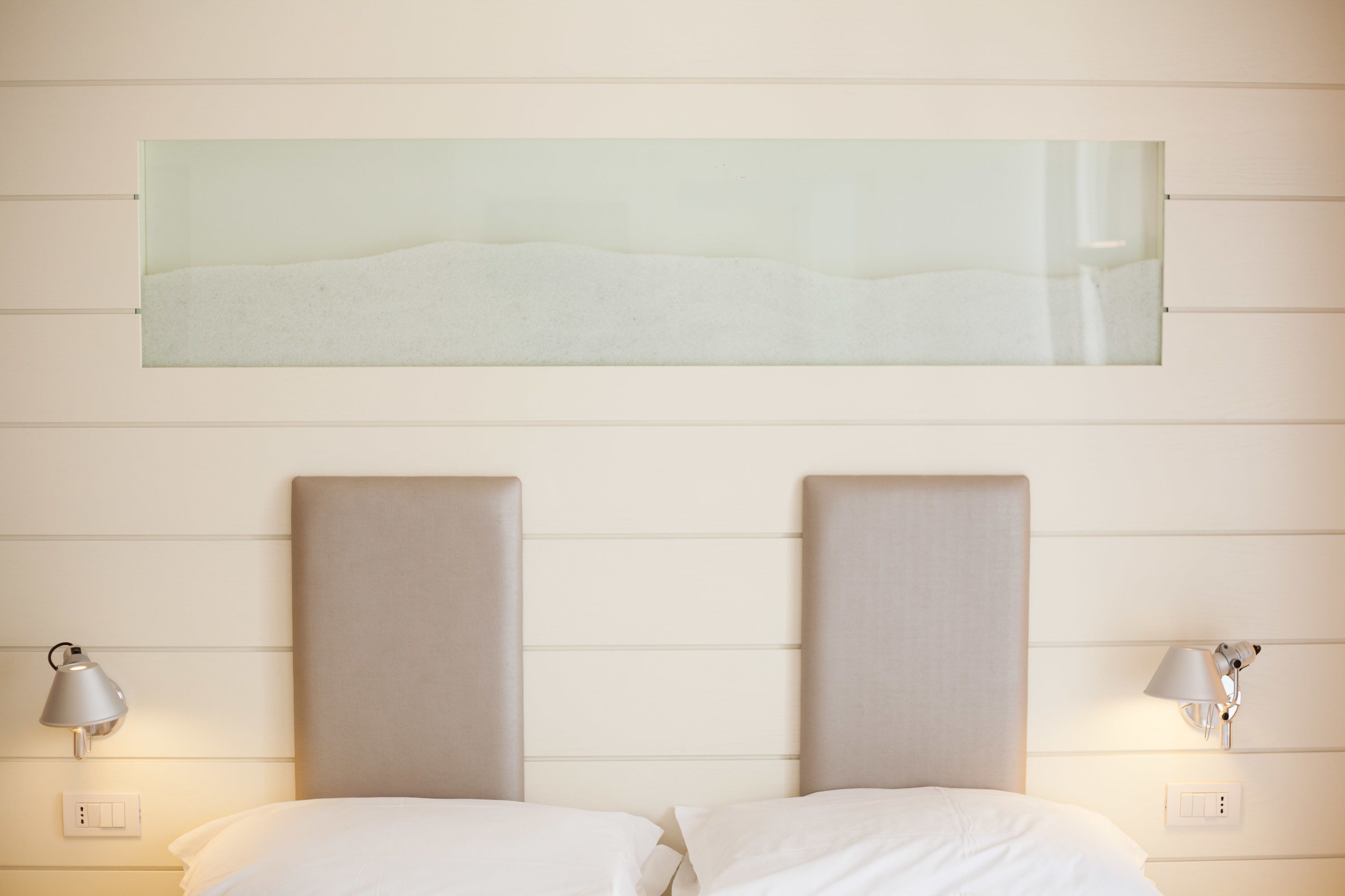 Quarto de hotel com uma cama grande, duas luminárias fixadas na parede e um espelho comprido sobre a cama.