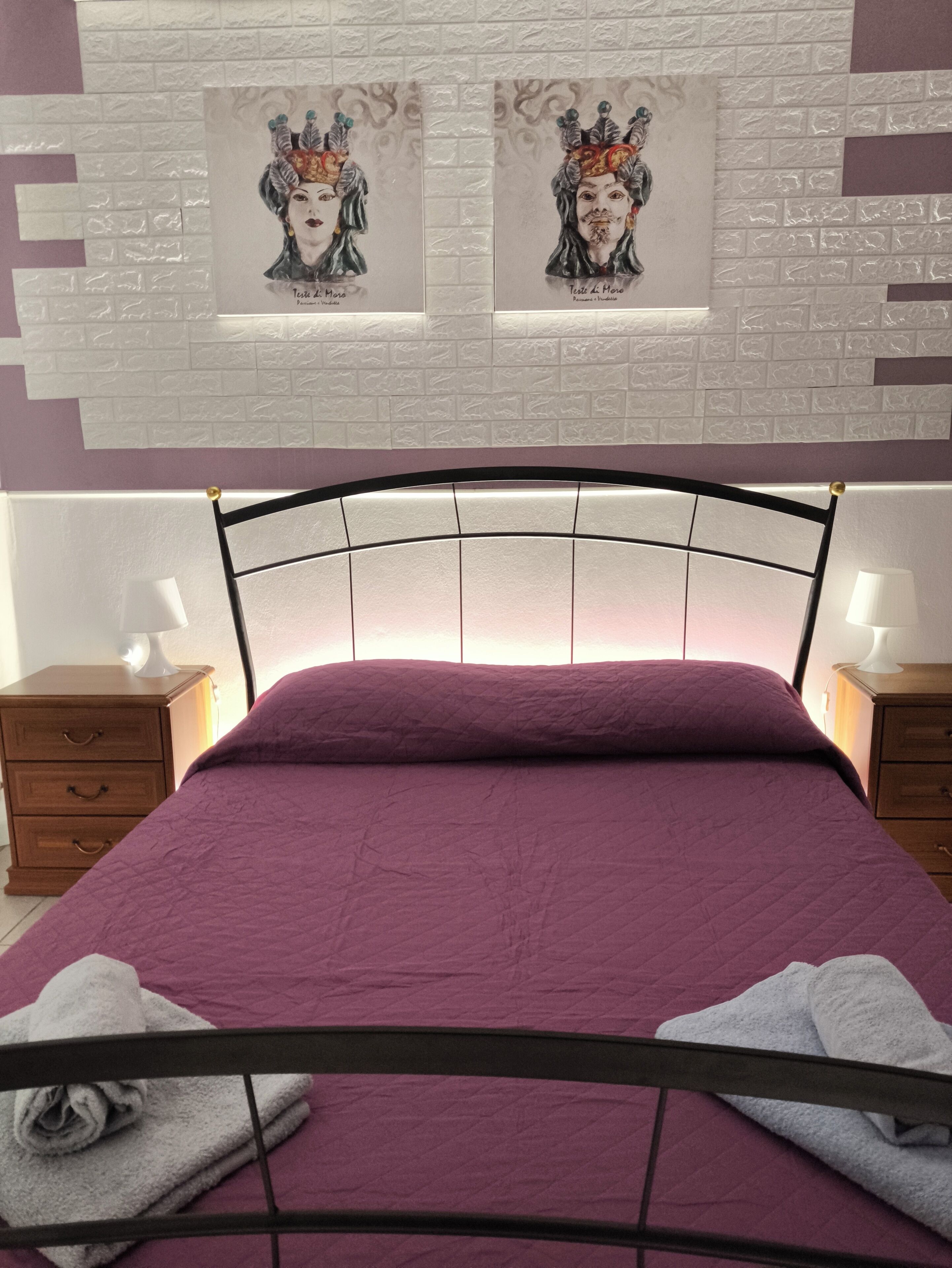 Quarto com cama, duas mesinhas de cabeceira, parede com padrão de tijolos brancos e duas obras de arte emolduradas acima da cama.