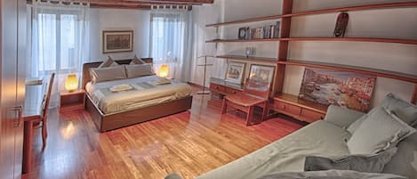 Quarto moderno com piso de madeira, cama com cabeceira, sofá, escrivaninha e prateleiras com livros e quadros.