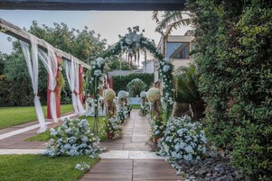 Uma passarela de madeira ladeada por flores brancas e vegetação, que conduz a uma estrutura vermelha com cortinas brancas.