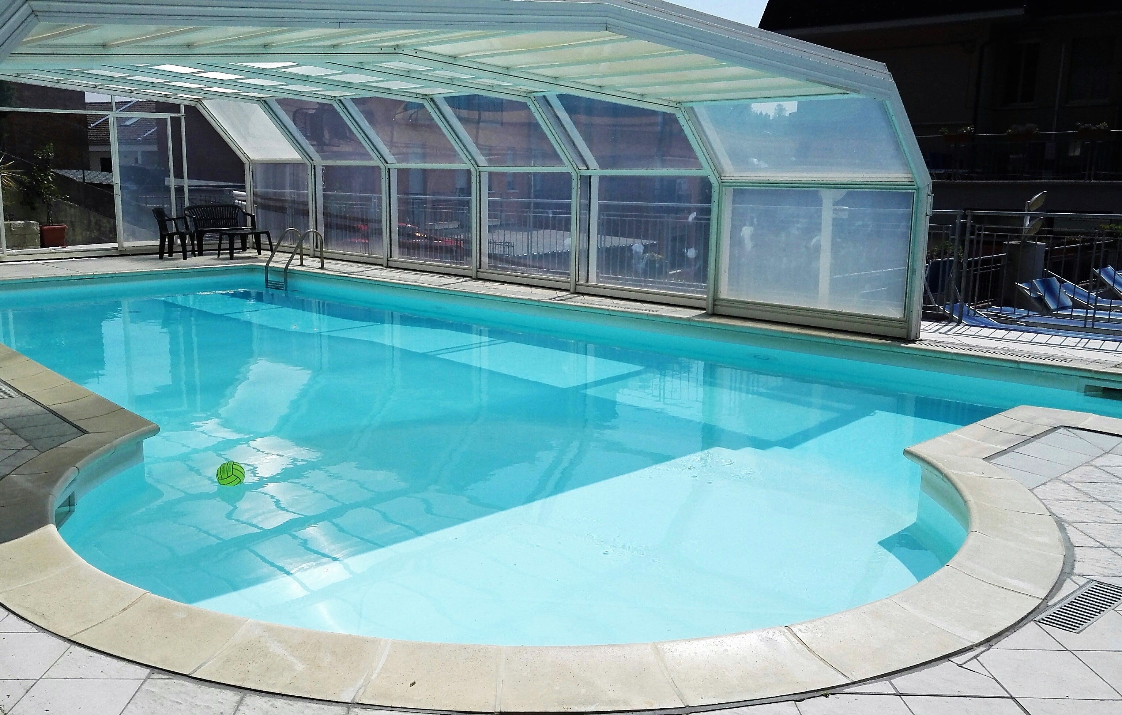 Piscina com cobertura de vidro, área de descanso com cadeiras e vista para os edifícios ao redor.