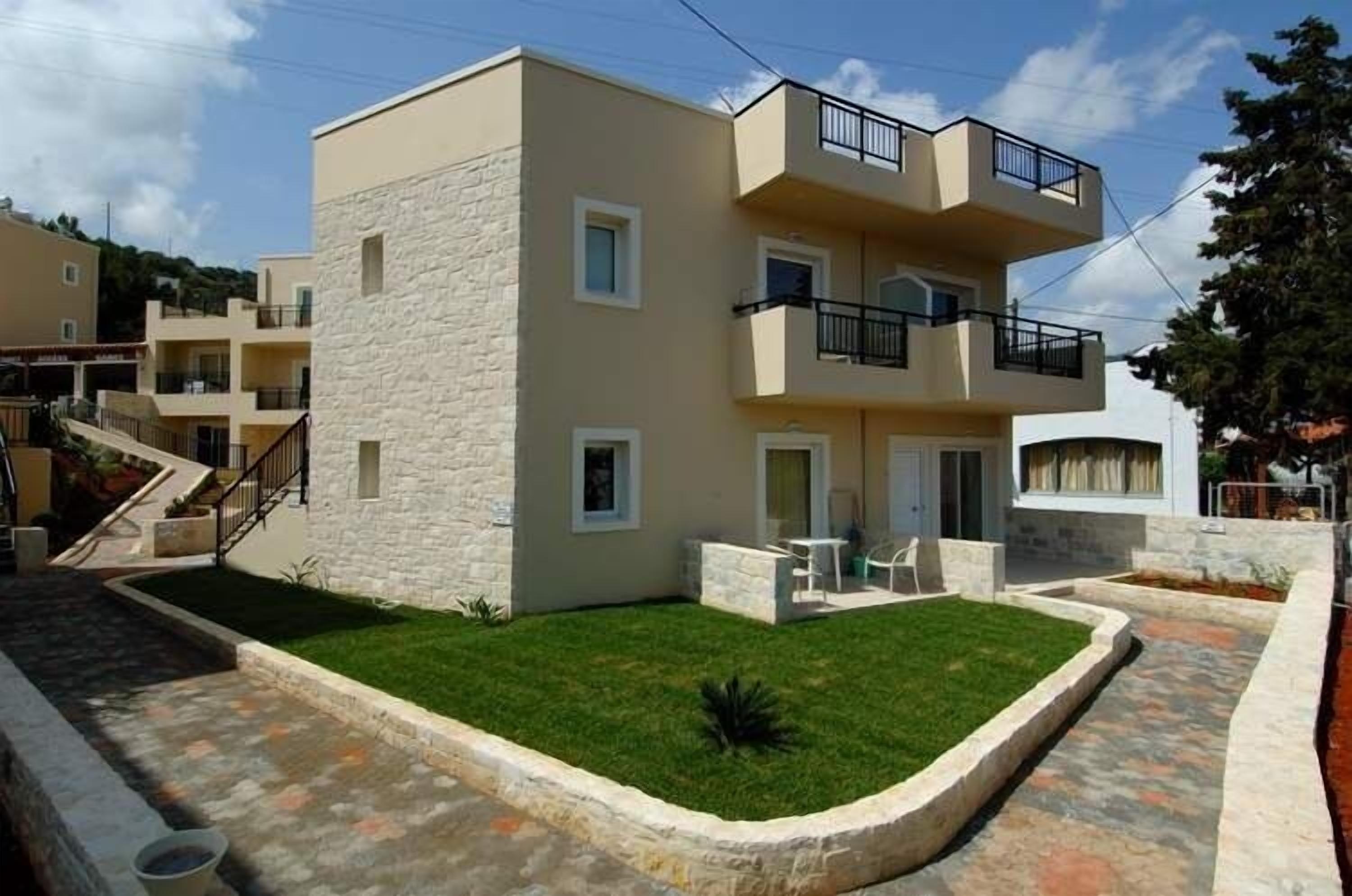 Um edifício residencial de vários andares com fachada de pedra, um pequeno jardim frontal com relvado e uma varanda com guarda-corpo preto.