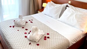 Uma cama bem arrumada com lençóis brancos e pétalas de rosa, cabeceira de madeira, mesa de cabeceira com abajur e quadro na parede.