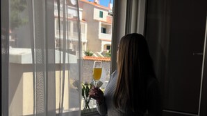 Uma pessoa segurando um copo com líquido amarelo perto de uma janela com cortinas brancas.
