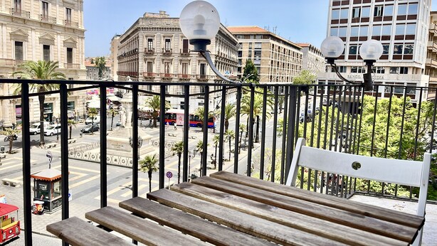 Varanda com banco de madeira, vista para uma rua movimentada com edifícios, palmeiras e uma estátua.