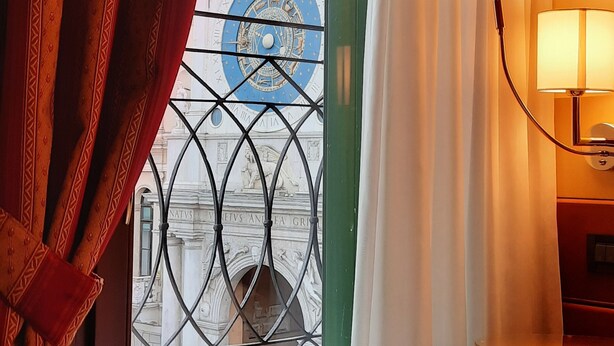 Quarto com janela, grade metálica decorativa e vista para um edifício com arquitetura clássica e um relógio azul.