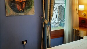 Quarto de hotel com cama, um quadro na parede, janela com cortinas e abajur em uma mesa de cabeceira.