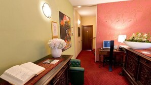 Uma área de recepção de hotel com balcão, um laptop, um vaso de flores e chão coberto por um tapete vermelho.