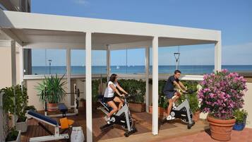 Uma área de ginástica coberta ao ar livre, com equipamentos de exercício, plantas em vasos e vista para o mar.