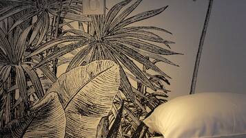 Quarto com cama, travesseiro branco, luminária pendente e um mural de palmeiras na parede.