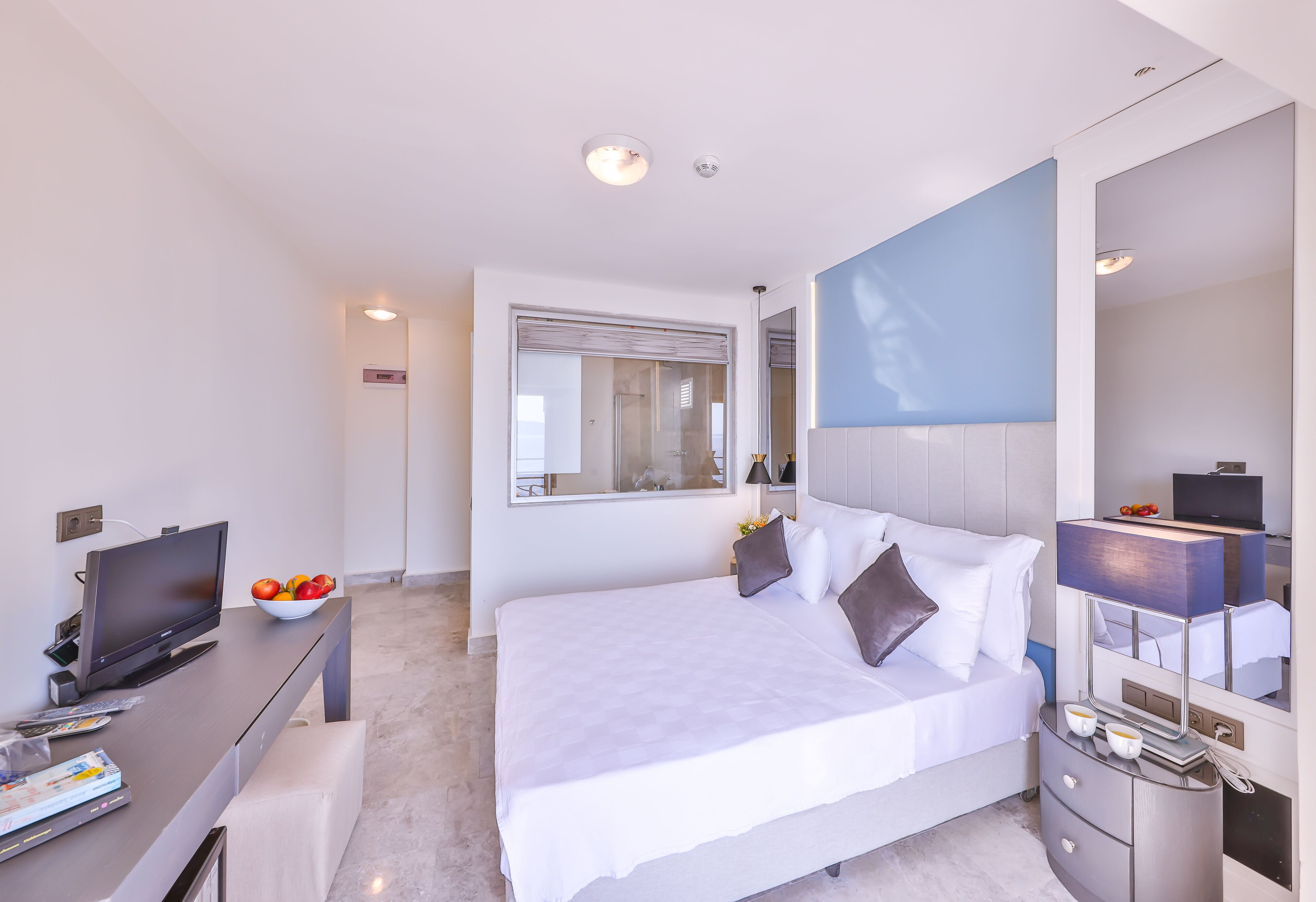Quarto de hotel moderno com uma cama grande, televisão de tela plana, escrivaninha com uma cesta de frutas e um espelho.