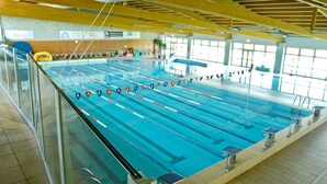 Uma grande piscina interior com várias pistas de natação, guarda-corpos de vidro e chão revestido de azulejos.