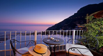 Uma varanda com vista para o mar e uma montanha ao entardecer. Há duas cadeiras e uma mesa com uma taça de vinho e um prato de comida.