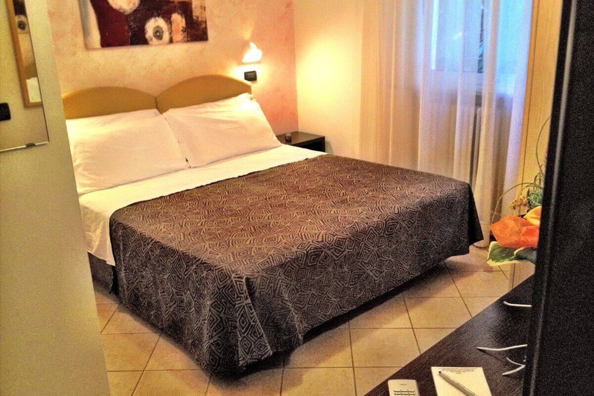 Quarto de hotel com cama, mesa de cabeceira, abajur e janela com cortinas.