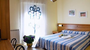 Quarto de hotel com cama, escrivaninha de madeira, cadeira, planta em vaso e janela com cortinas.