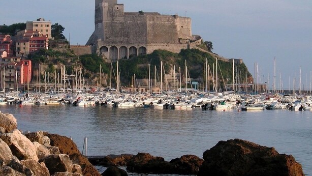 Uma vila costeira com um castelo histórico numa colina, uma marina com muitos barcos, e arribas rochosas em primeiro plano.