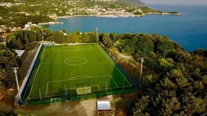 Vista aérea de um campo de futebol cercado por árvores densas e uma paisagem costeira com colinas e edifícios ao fundo.