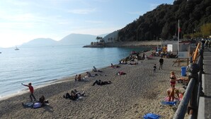Uma praia com pessoas tomando sol e nadando, uma torre de salva-vidas e uma vista clara do mar e montanhas ao longe.