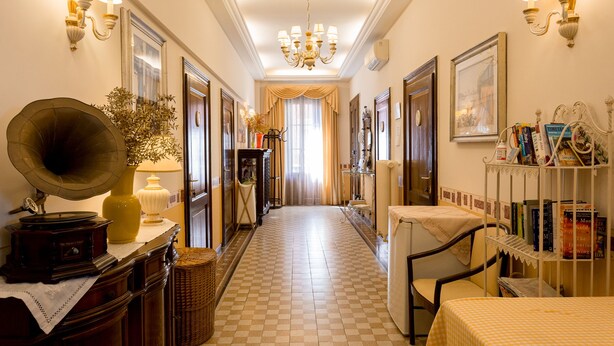 Um corredor de hotel com piso xadrez, um candelabro, arandelas, um gramofone antigo e uma estante de livros.