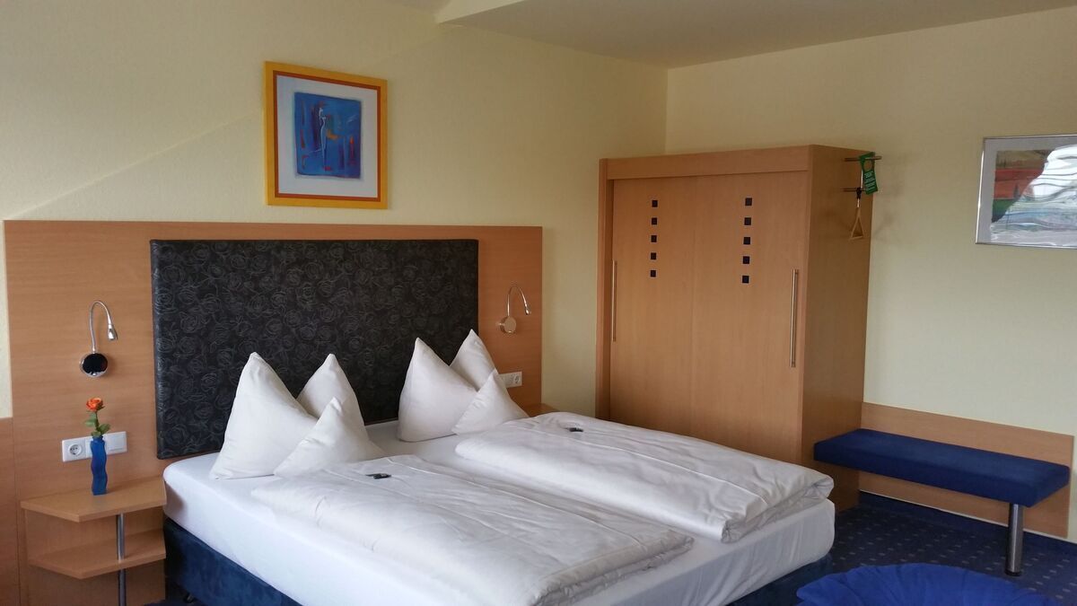 Quarto de hotel com uma cama grande, um guarda-roupa de madeira, um banco e duas molduras na parede.