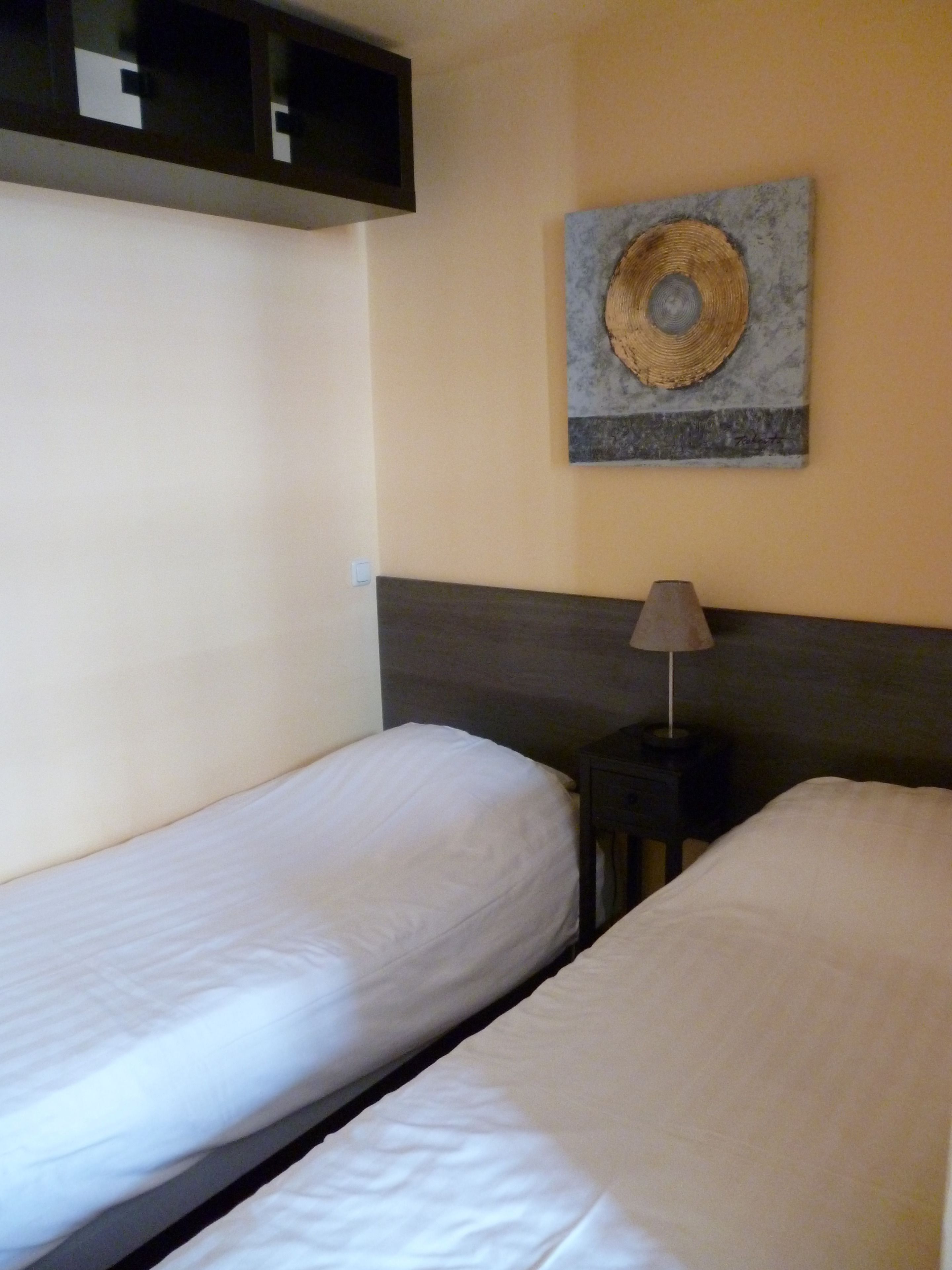 Quarto de hotel com duas camas de solteiro, mesa de cabeceira com abajur, prateleira na parede e um objeto decorativo na parede.