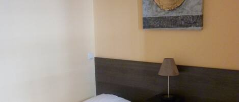 Quarto de hotel com duas camas de solteiro, mesa de cabeceira com abajur, prateleira na parede e um objeto decorativo na parede.