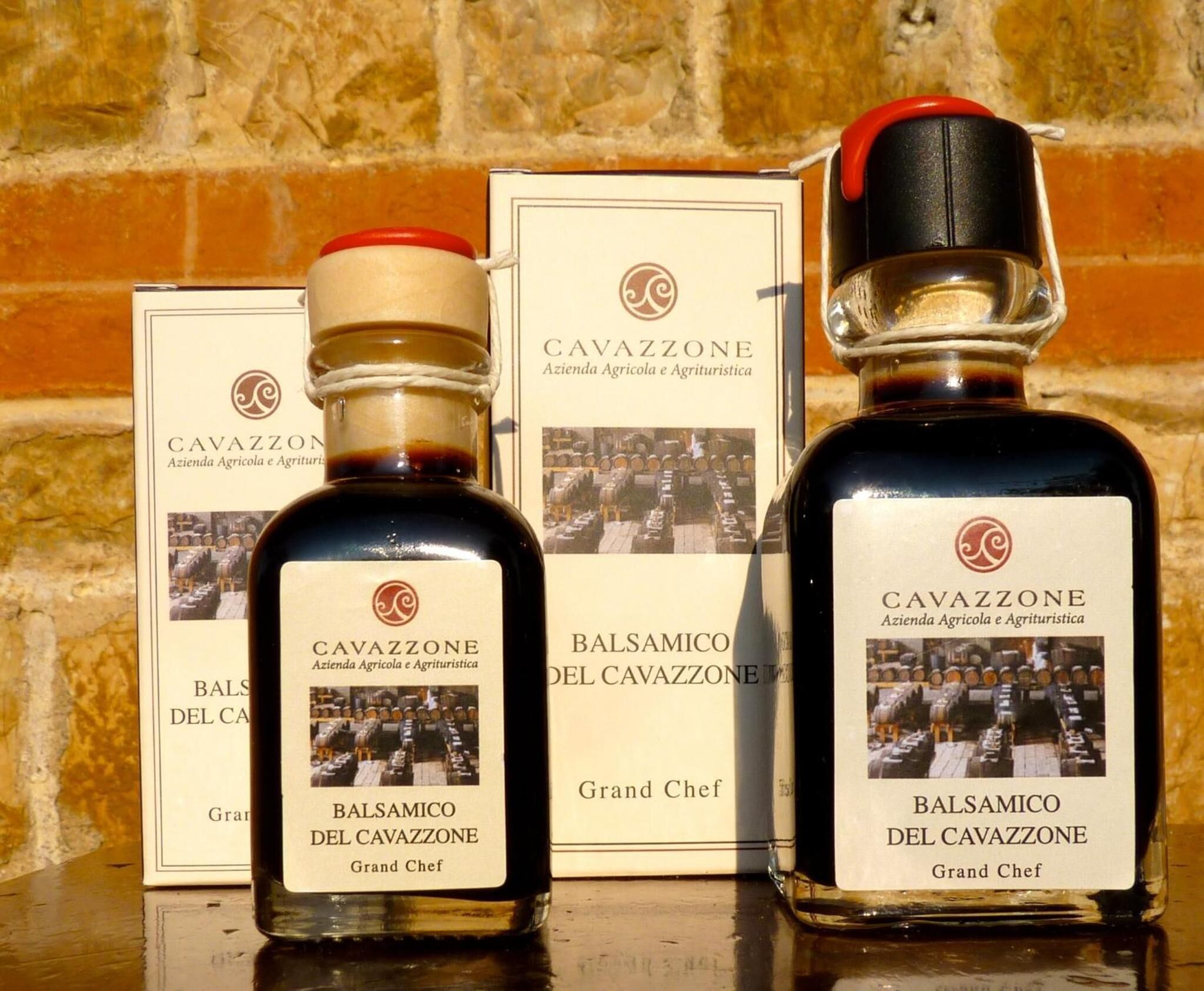 Três garrafas de vinho CAVAZZONE Balsamico Del Cavazzone Grand Chef, juntamente com as suas caixas, apoiadas numa parede de tijolos.