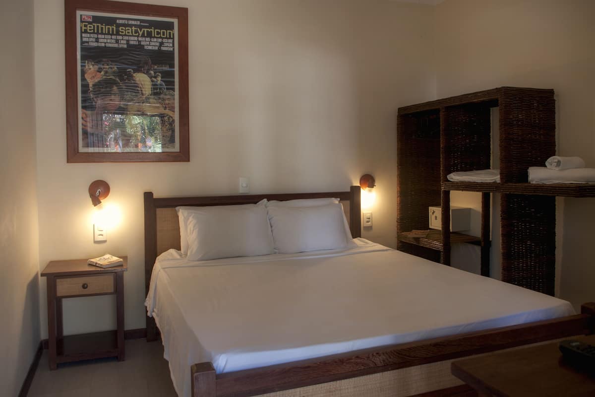 Uma cama bem arrumada com roupa de cama branca, uma pequena mesa de cabeceira com abajur e um cartaz na parede.
