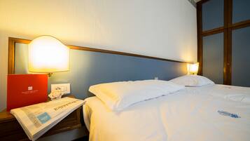 Quarto de hotel com uma cama bem arrumada, uma pasta vermelha na mesa de cabeceira e uma luminária iluminando o ambiente.