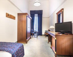 Quarto de hotel com cama, escrivaninha com televisão, poltrona, guarda-roupa e janela com cortinas.