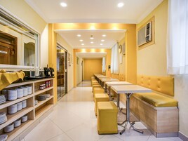 Corredor de hotel com uma fileira de mesas e bancos, área de serviço com equipamentos de cozinha e um espelho na parede.