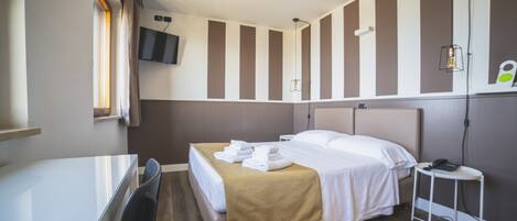 Quarto de hotel com cama, escrivaninha, cadeira, televisão presa na parede e mesa de cabeceira com telefone.