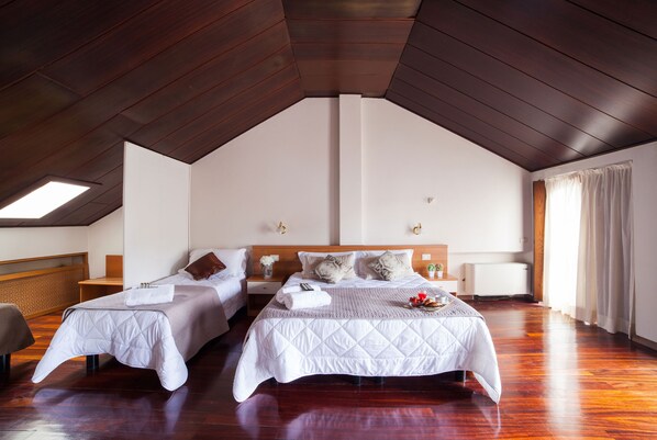 Quarto de hotel com duas camas, piso de madeira, teto inclinado e uma janela grande com cortinas.