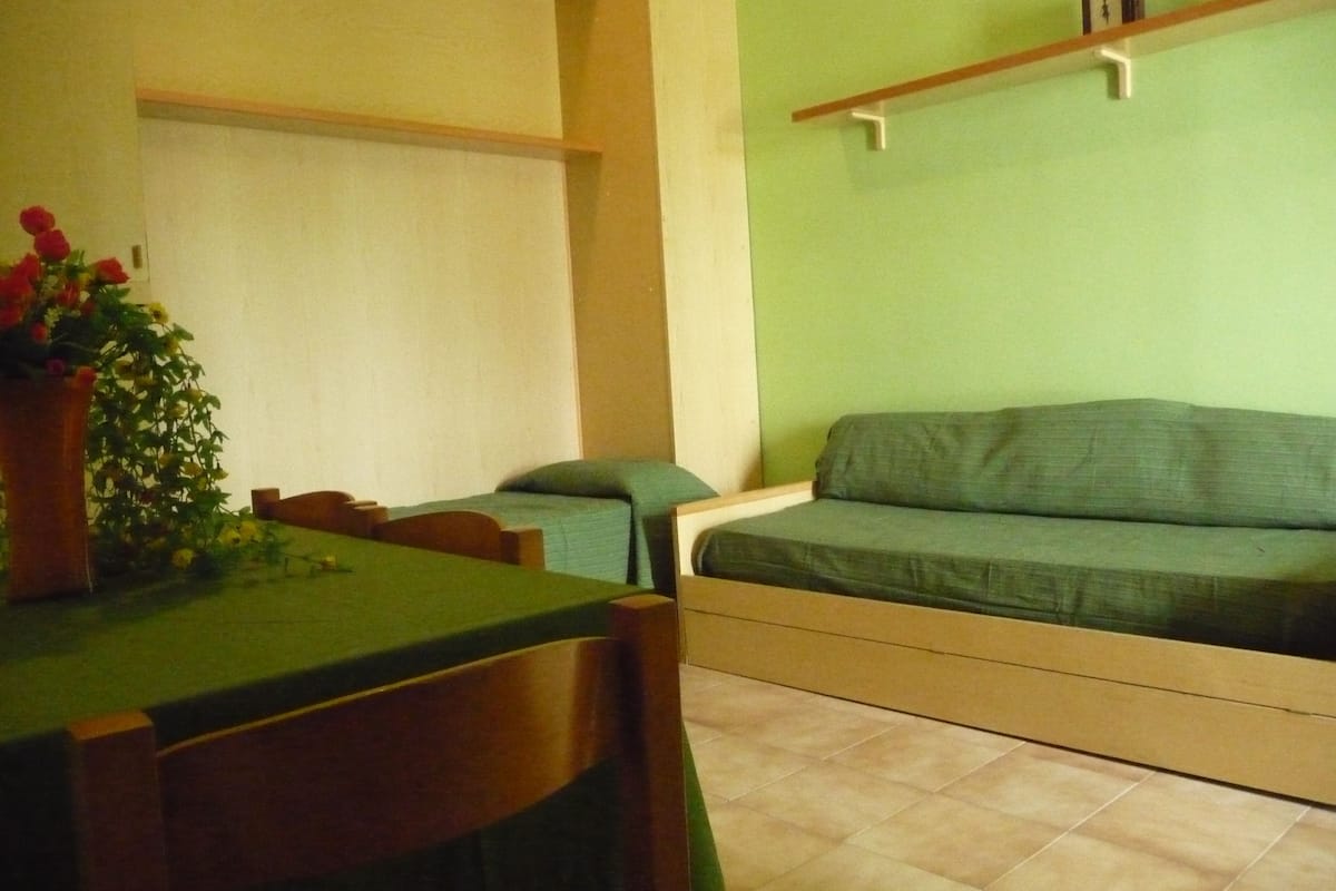 Quarto pequeno com sofá verde, mesa de madeira e uma cadeira. Há uma pequena planta sobre a mesa.