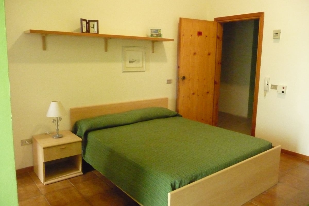 Quarto com cama, mesa de cabeceira, prateleira com fotos, porta e tomada na parede.