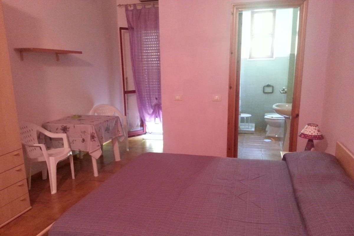 Quarto com cama, uma mesinha e cadeira, cômoda e banheiro visível por uma porta aberta.