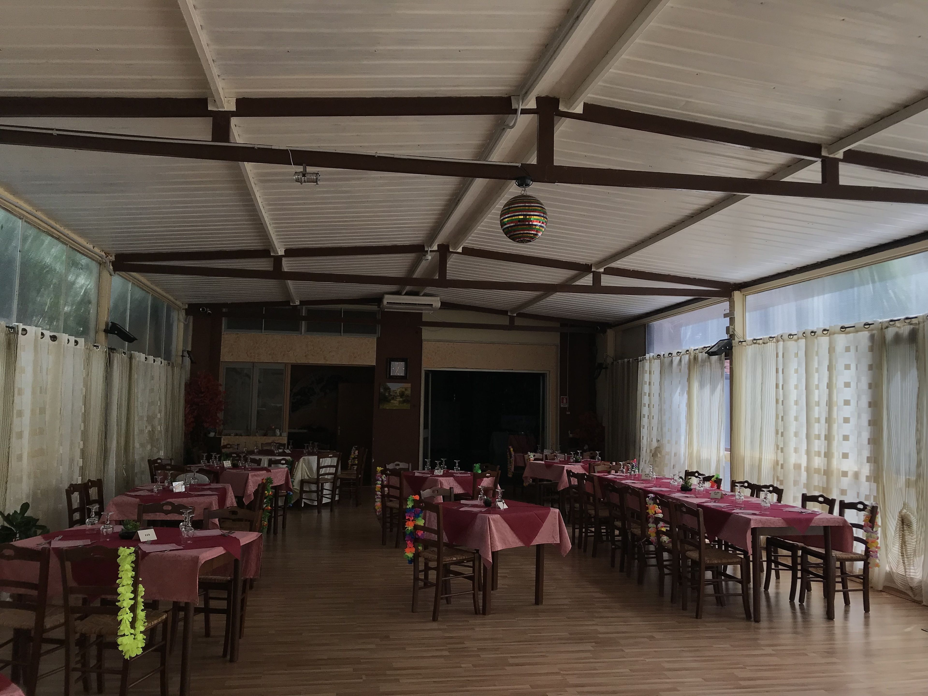 Um salão amplo com várias mesas e cadeiras, teto alto com vigas de metal e grandes janelas com cortinas.