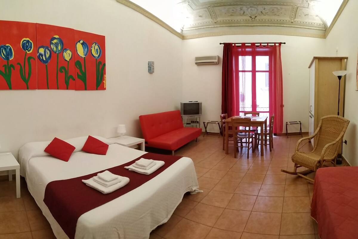 Quarto de hotel com cama, sofá vermelho, mesa de jantar com cadeiras, televisão e pintura de flores na parede.