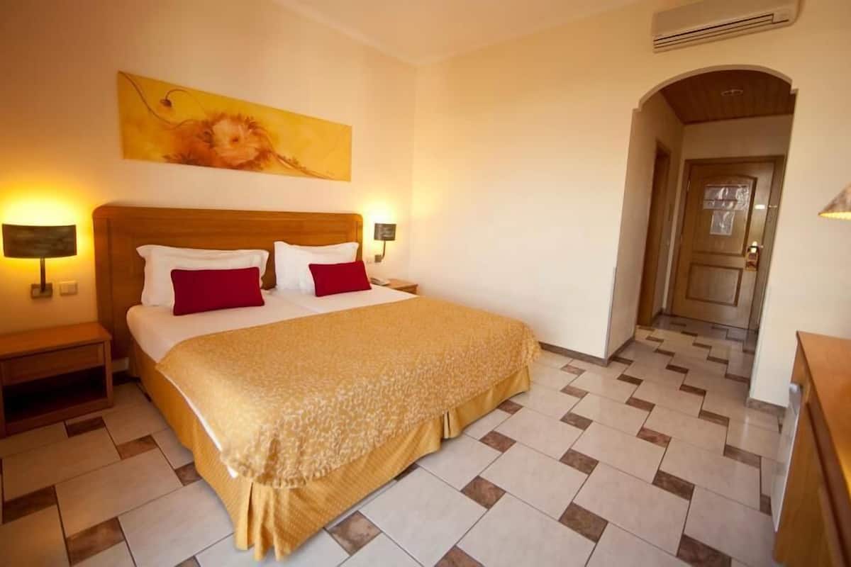 Quarto de hotel com uma cama grande, duas mesas de cabeceira com luminárias, uma obra de arte na parede e piso ladrilhado.